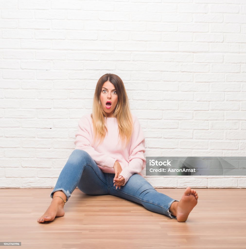 Young Adult Woman Sitting On The Floor Over White Brick Wall Afraid And