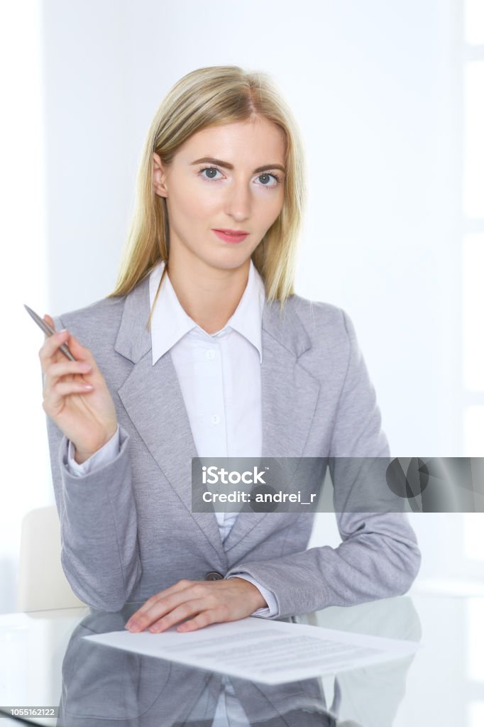 Blonde Business Woman Examining Contract While Sitting At The Glass Desk In Office Background