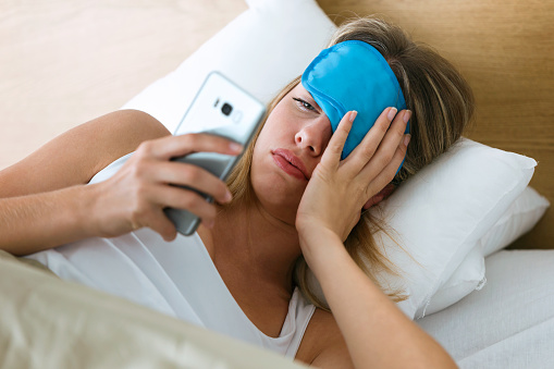 Tired Young Woman Lying In Bed Pulling Up Sleeping Mask To See What