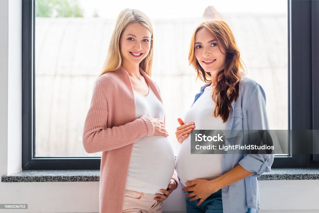 Smiling Pregnant Women Standing Near Window And Looking At Camera Stock