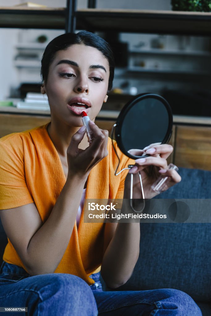 Beautiful Mixed Race Girl In Orange Shirt Applying Red Lipstick At Home