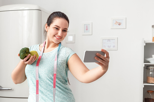 Body Care Chubby Girl Standing With Tape Measure Around Neck In Kitchen Taking Selfie On