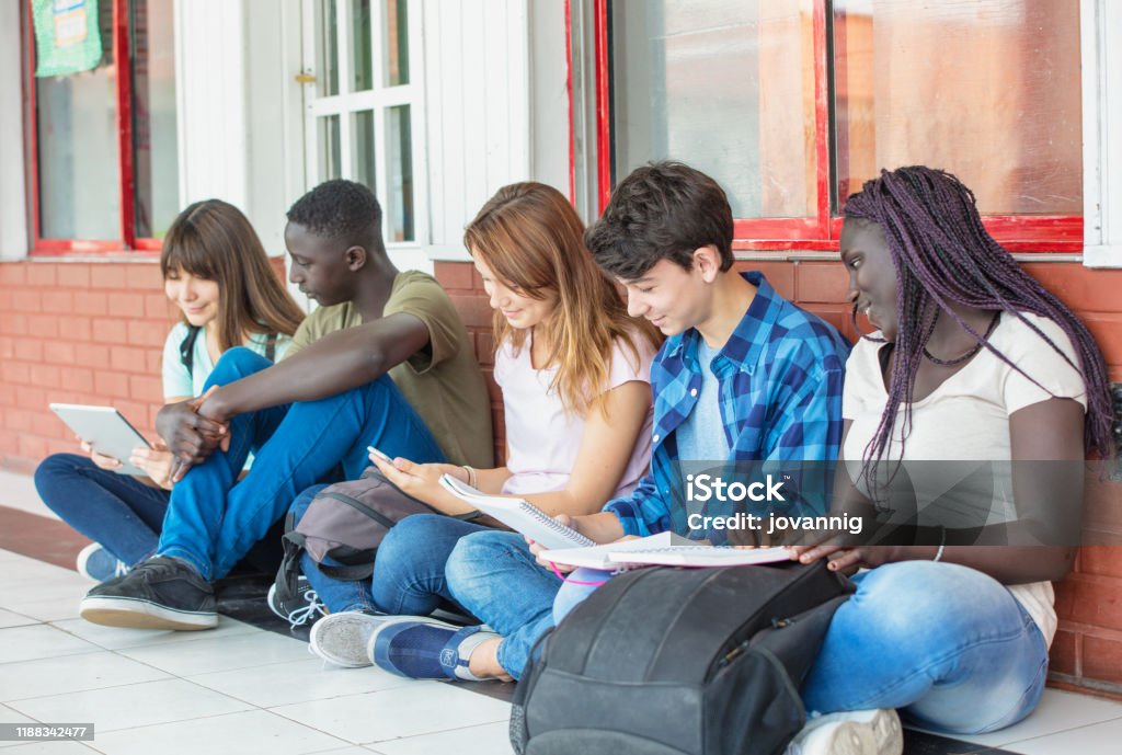 Multi Race Classroom Using Books And Tablets Seated In Schoolyard Stock