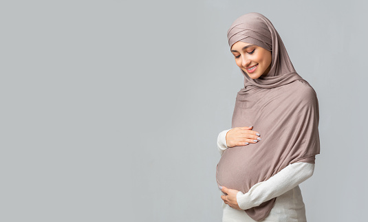 Pregnant Muslim Woman Embracing Her Belly Posing Over Grey Background