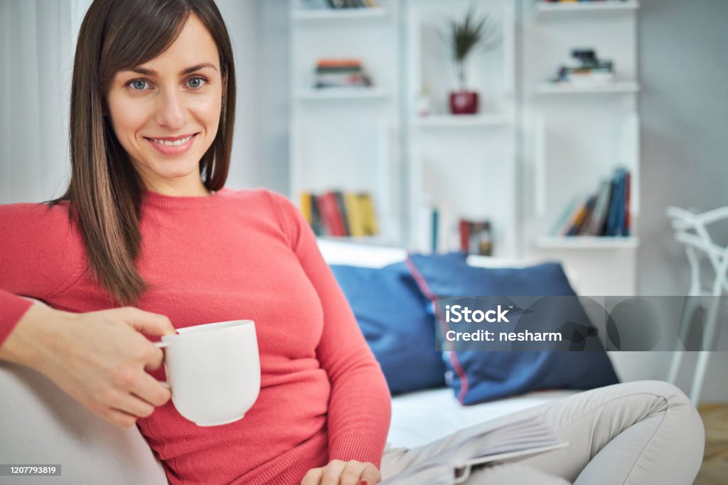 Attractive Brunette Sitting On Sofa With Crossed Legs Reading Book And