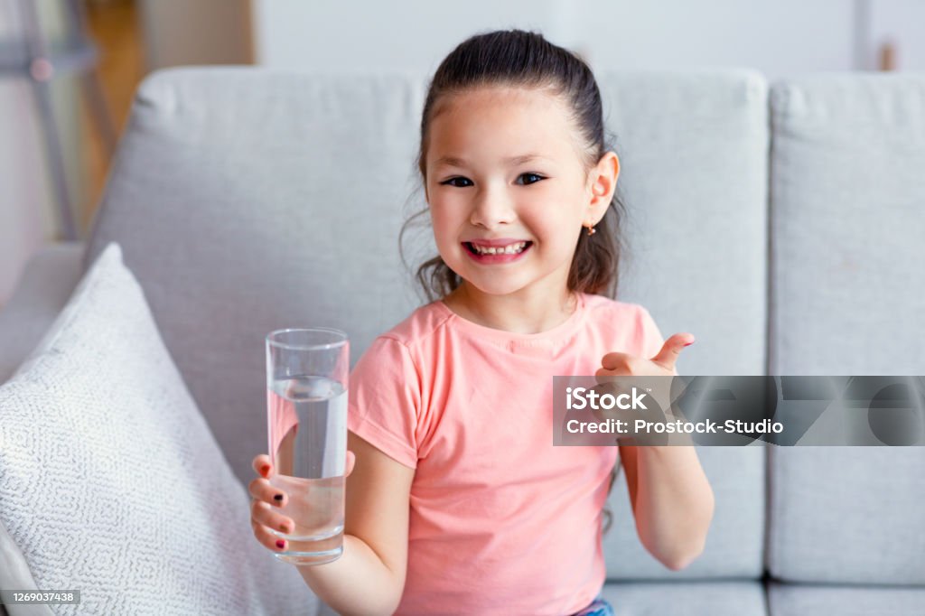 Chinese Girl Holding Glass Of Water Gesturing Thumbsup Sitting Indoors