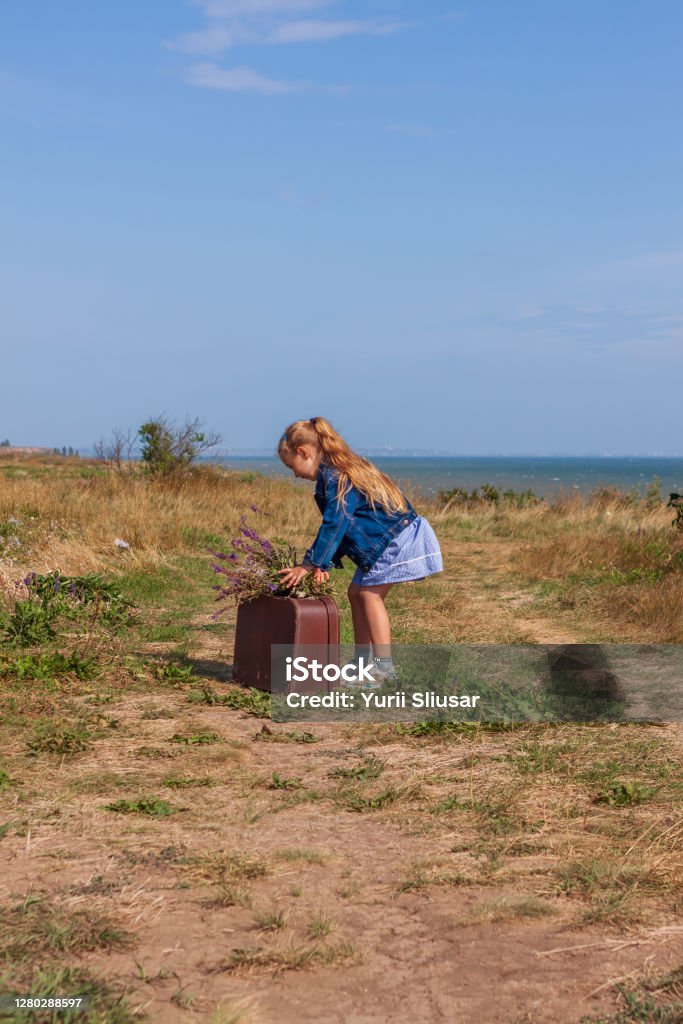 Little Blonde Girl In Denim Jacket Blue Dress With Vintage Suitcase And Flowers Bouquet Offroad