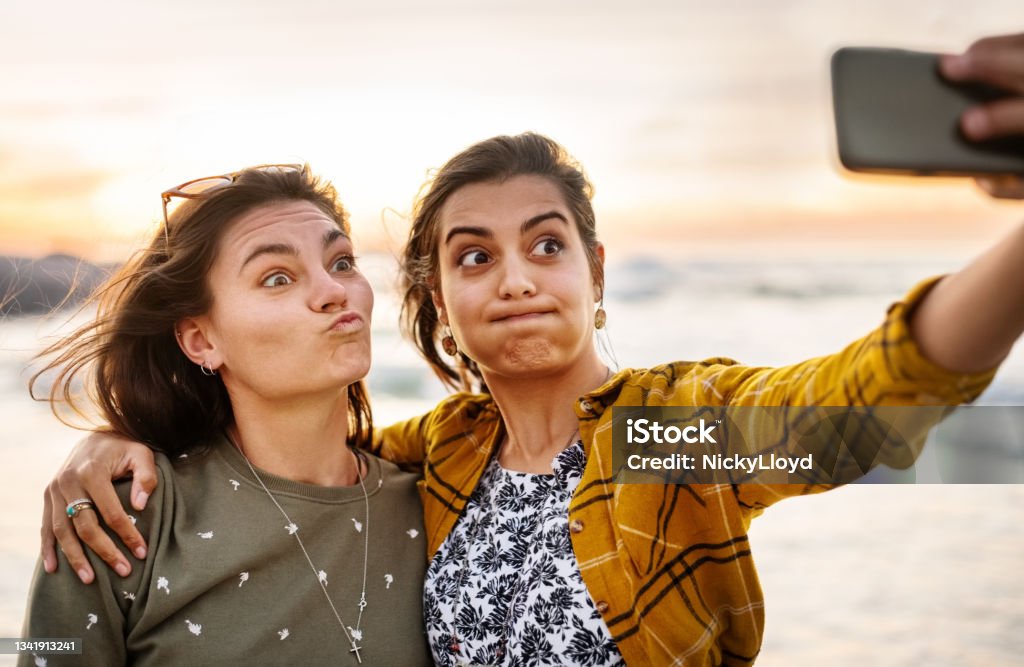 Female Friends Making Funny Faces While Taking Selfies At The Beach