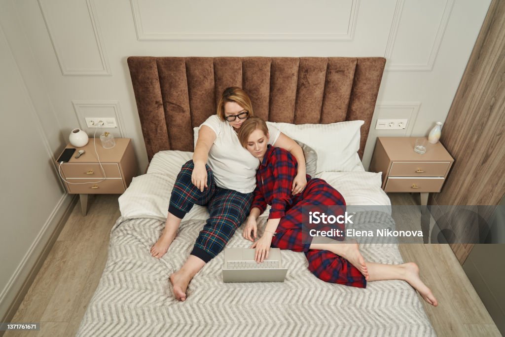 Lesbian Couple In Pajamas Lies On The Bed In Front Of Them Open Laptop