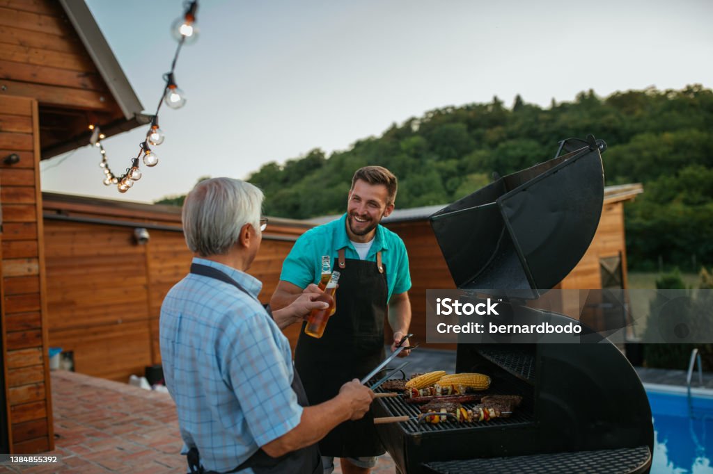 Young Adult Man And His Father Toasting With Beer While Barbecuing In