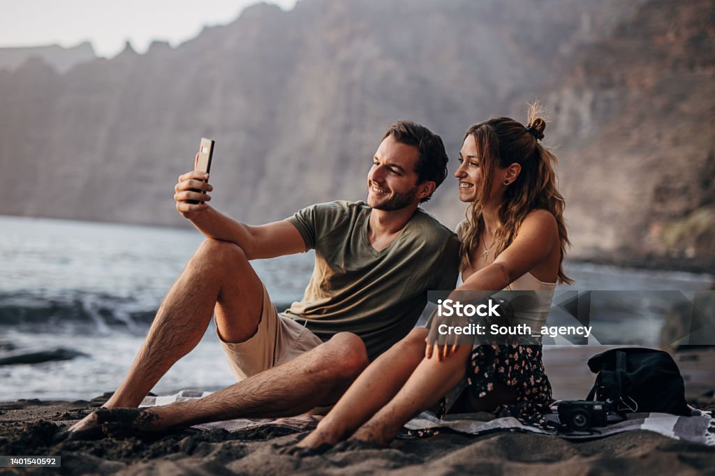 Young Couple Sitting On The Beach At Sunset And Taking A Selfie Stock