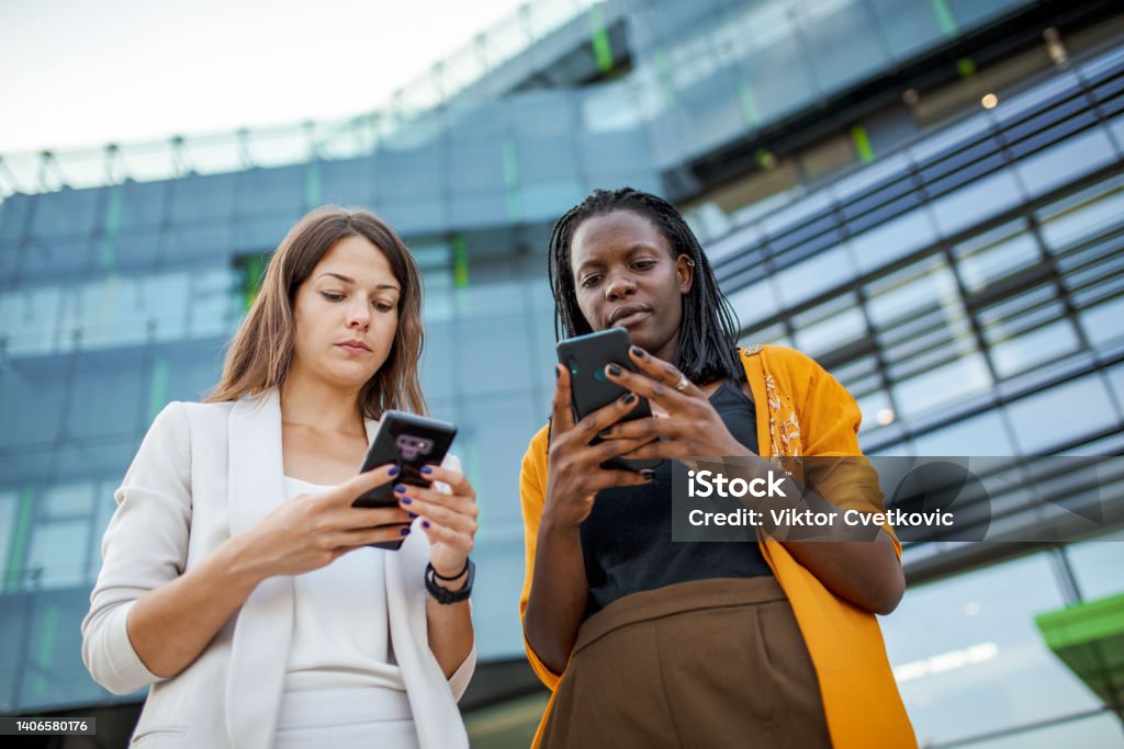 Two Multiracial Business Women Using Their Mobile Phones In Front Of An ... Two Multiracial Business Women Using Their Mobile Phones In Front Of An ...