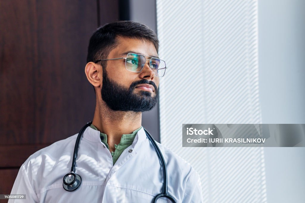 Portrait Of Male Indian Doctor Wearing White Coat In Clinic Office