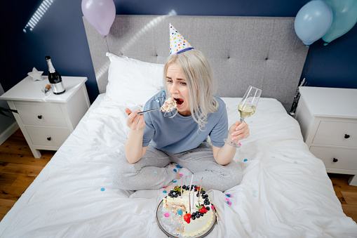Sad Upset Woman In Pajama And Party Cap Drinking Champagne And Eating