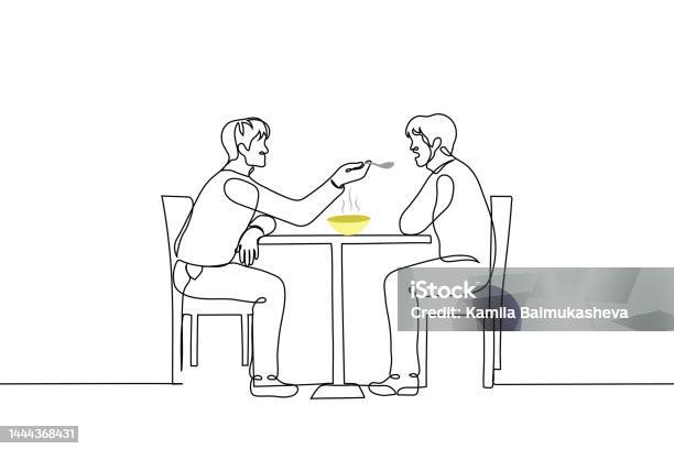 Man Sits At A Table And Feeds From A Spoon Another Man Sitting Opposite
