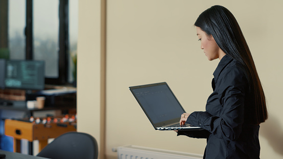 Asian Software Developer Coming At Desk And Sitting Down Holding Laptop With Coding Interface
