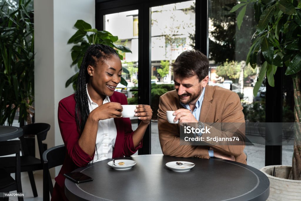 Young Man And African American Woman Sitting In Cafeteria Drinking