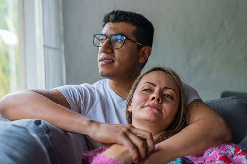Latin Couple Both Are Sitting On The Sofa In The Living Room Of Their House Hugging Enjoying A