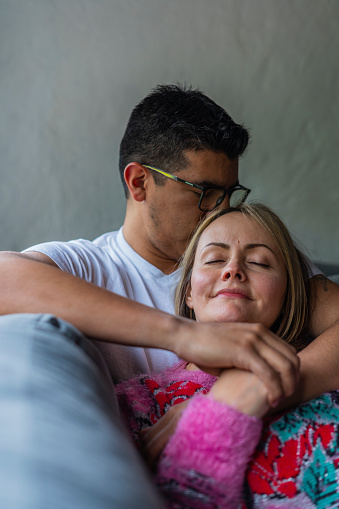 Latin Couple Both Are Sitting On The Sofa In The Living Room Of Their House Hugging Enjoying A