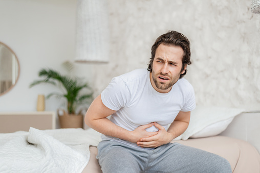 Unhappy Millennial Caucasian Guy With Stubble Wake Up Sits On White Bed