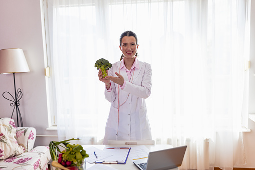 Female Nutritionist Holding Broccoli And Standing In Her Clinic Stock