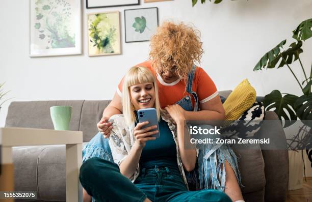 Smiling Lesbian Couple Sit On The Sofa Embracing Each Other And Using