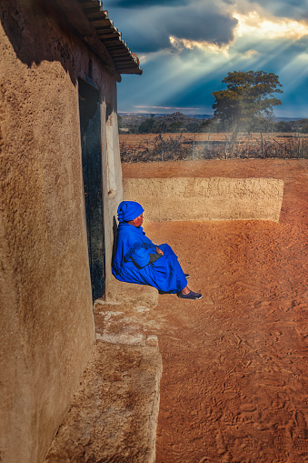 African Old Woman In A Blue Dress Sited In Front Of Her Home In The Sandy Yard In A Late