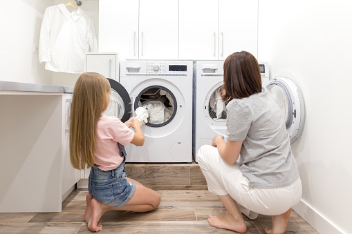 Mother And Daughter Helper In Laundry Room Near Washing Machine And