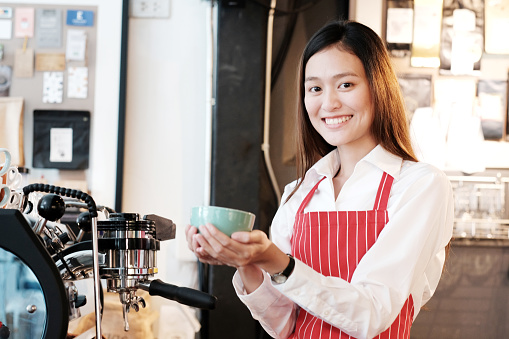 젊은 아시아 여성 바리 스타 들고 커피 컵 카페 카운터 배경 중소 기업 소유자 음식 및 음료 산업 개념에 얼굴을 미소와 기계류에
