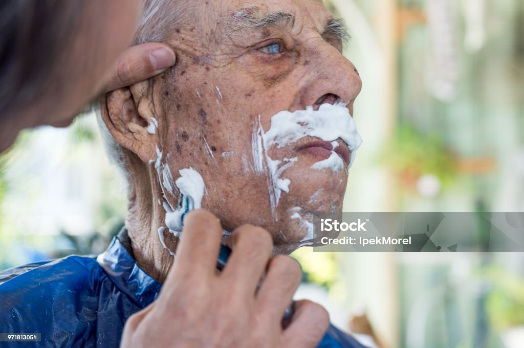 Old Man Getting His Beard Shaved By Young Skilled Man At Home Stock